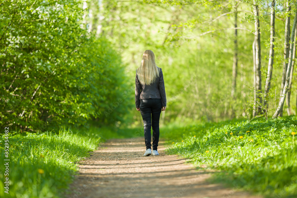 One young blonde woman slowly walking on natural trail in green forest ...