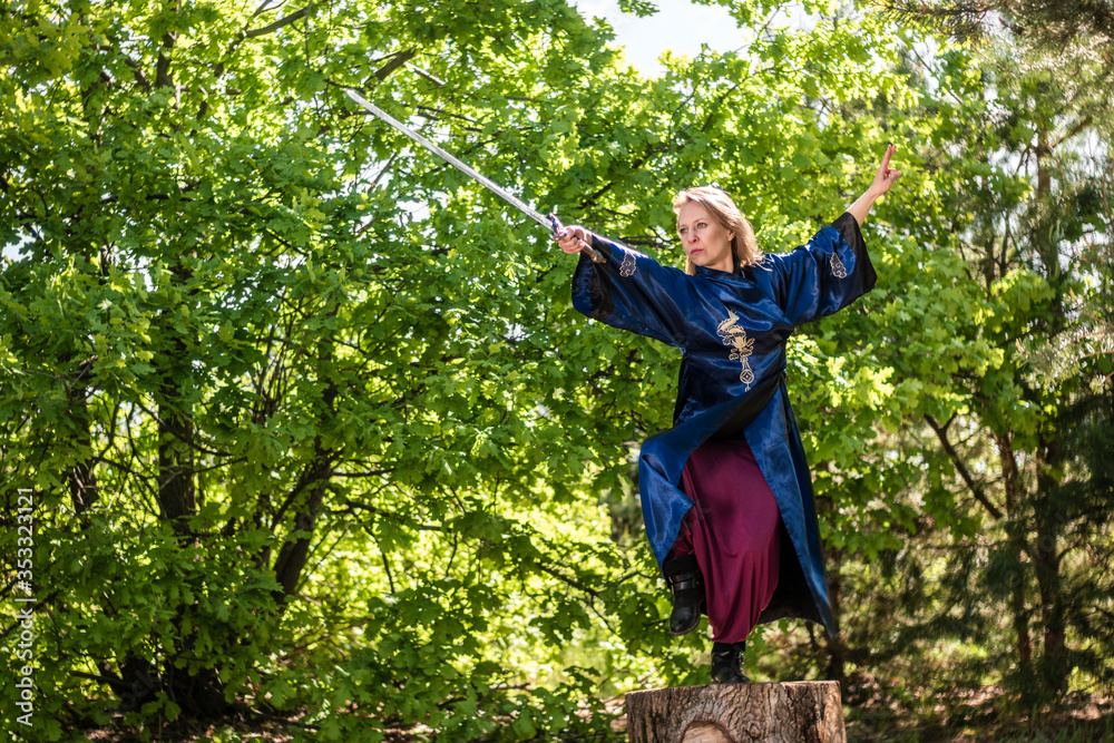 A female warrior with a sword poses on a stump in the forest. Wushu ...