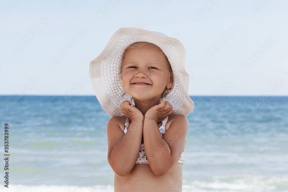 Little girl in a white beach hat and bikini by the sea put both hands ...