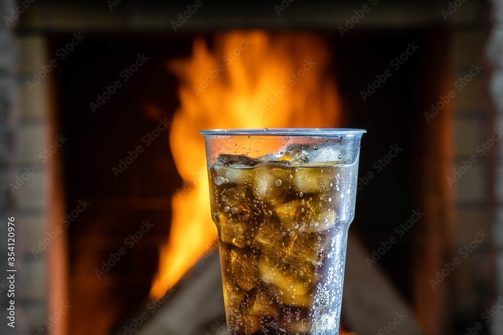 Plastic Glass of cola with ice in front of burning fireplace in a ...