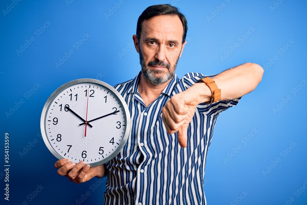 Middle age hoary man holding big clock standing over isolated blue ...