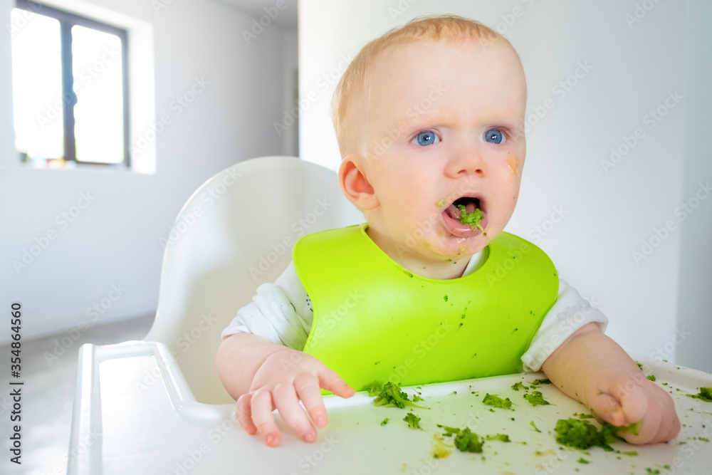 Annoyed excited baby eating broccoli vegs, looking away with open mouth. Little child wearing ...