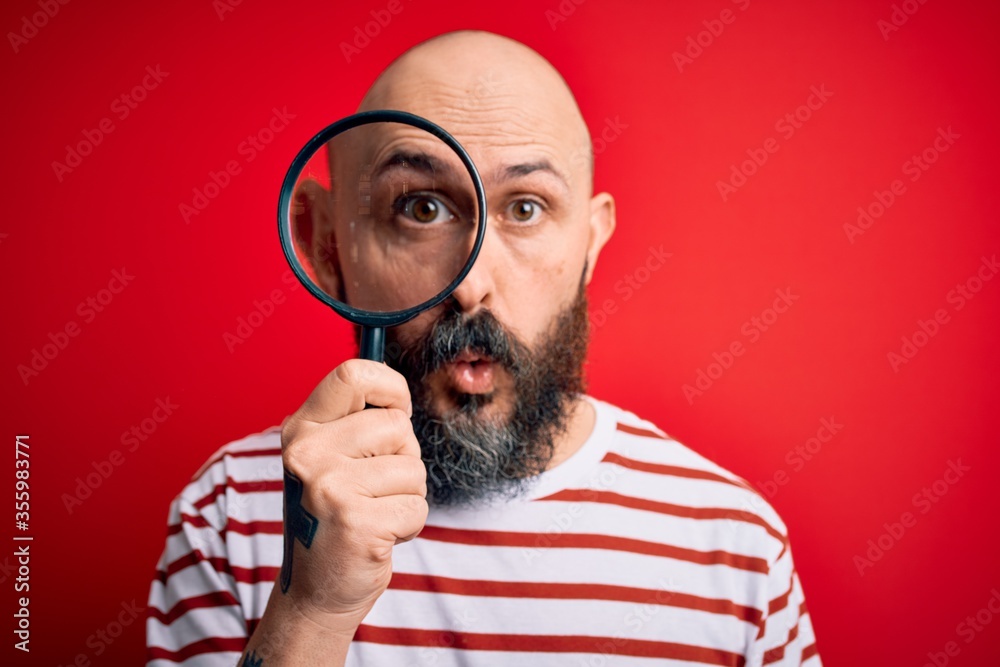 Handsome detective bald man with beard using magnifying glass over red ...