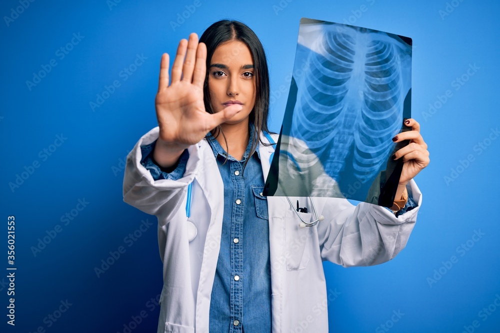 Young beautiful brunette doctor woman wearing stethoscope holding chest ...