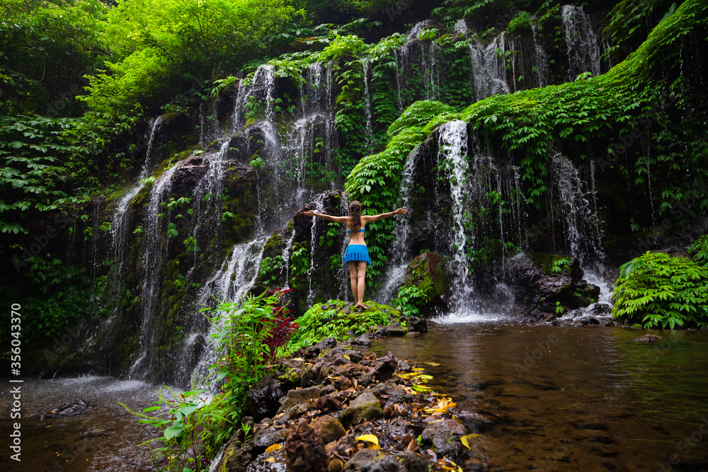 Excited Caucasian woman raising arms in front of waterfall. View from ...