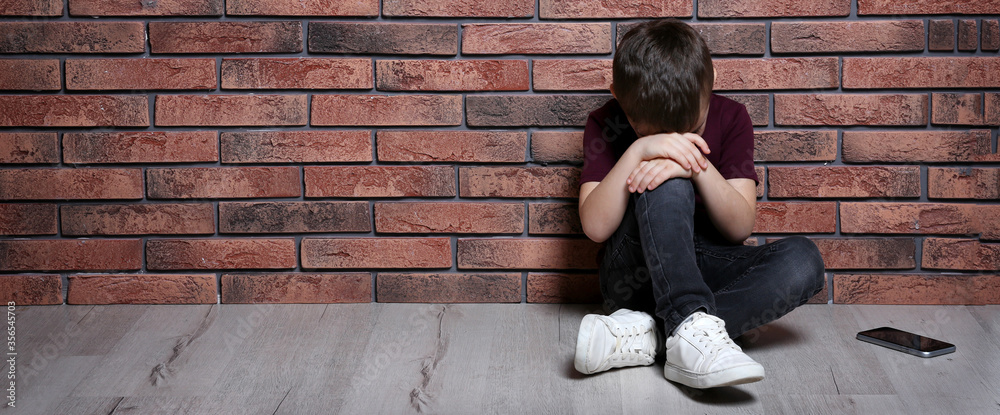 Sad little boy with mobile phone sitting on floor near brick wall ...