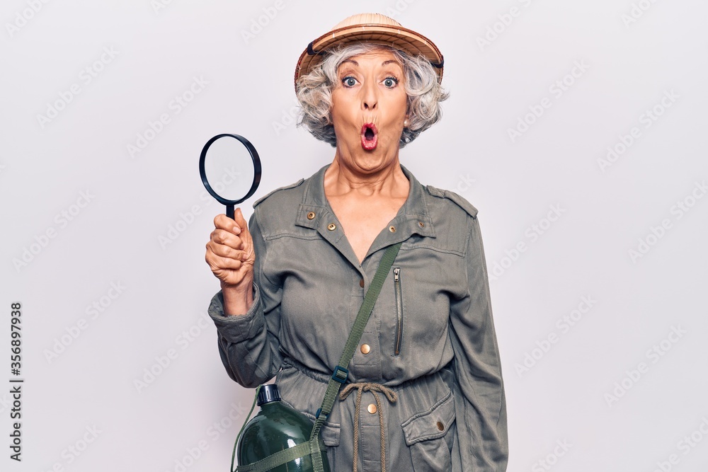 Senior grey-haired woman wearing explorer hat holding magnifying glass ...