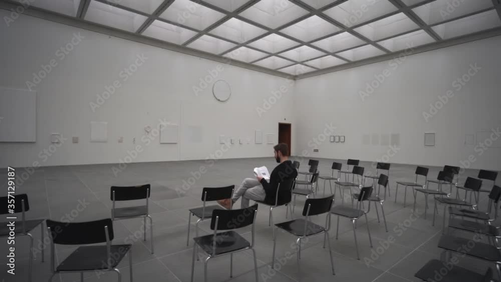 Lonely young man sits on one of the many chairs in a bare white museum ...