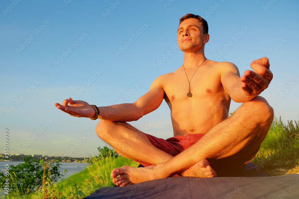 Fit muscular man doing yoga by the water sitting in a lotus pose with ...