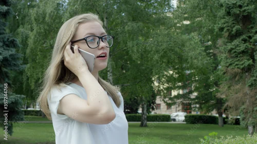 Blonde bespectacled girl talking on the phone while standing in park ...