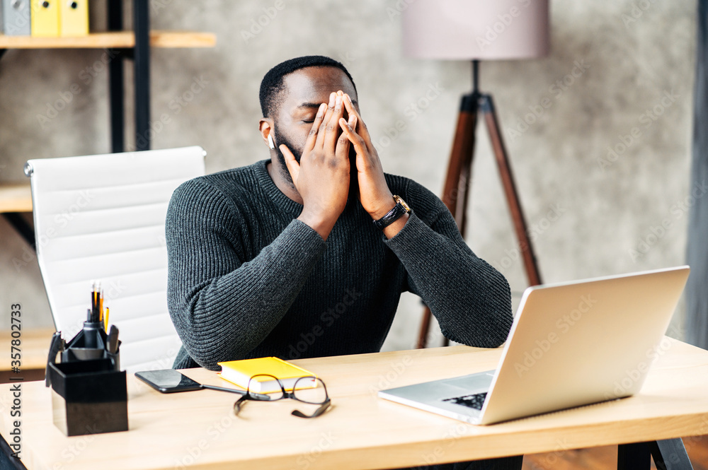 Exhausted African-American office employee sits at the desk with a ...
