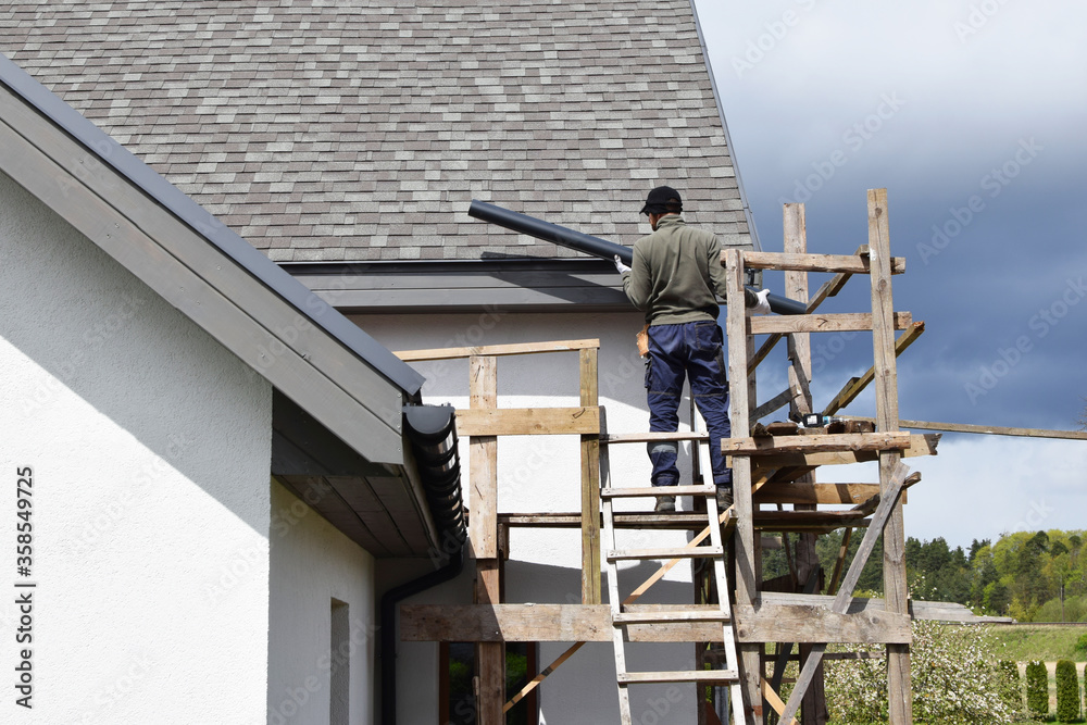 Construction worker installing plastic roof rain gutter gray shingles ...