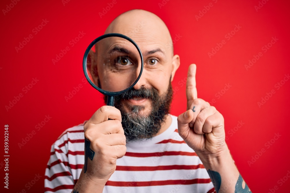 Handsome detective bald man with beard using magnifying glass over red ...