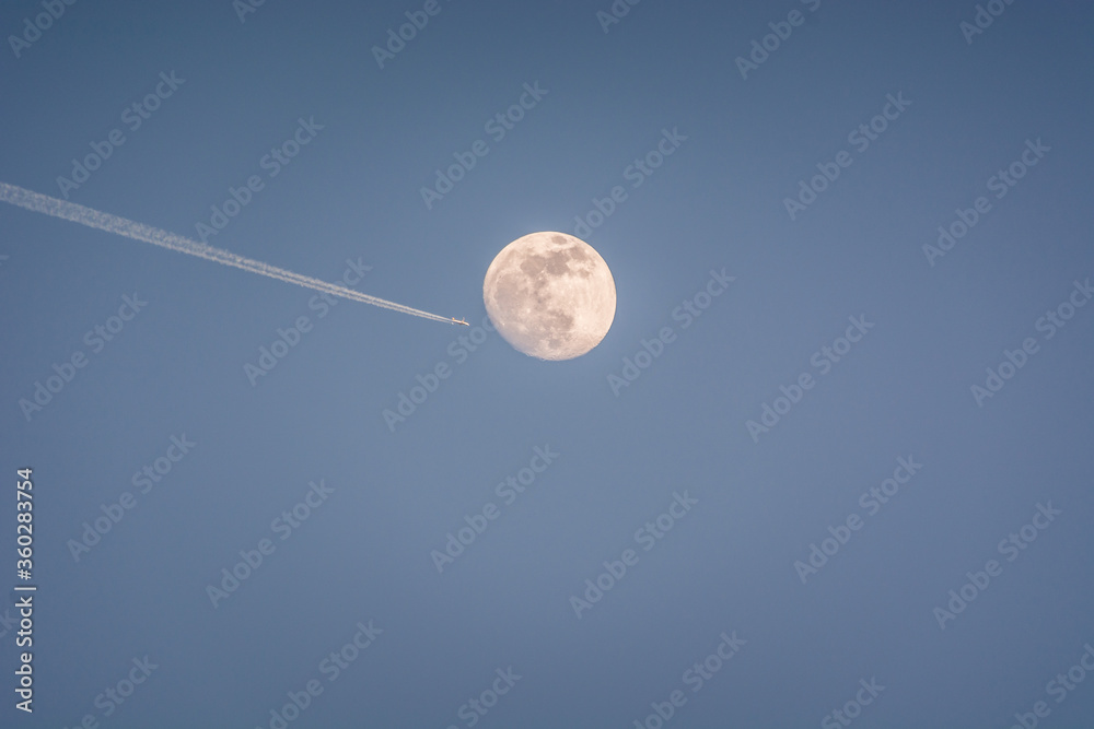 Airplane leaves a trail while passing a full moon on a clear blue sky ...