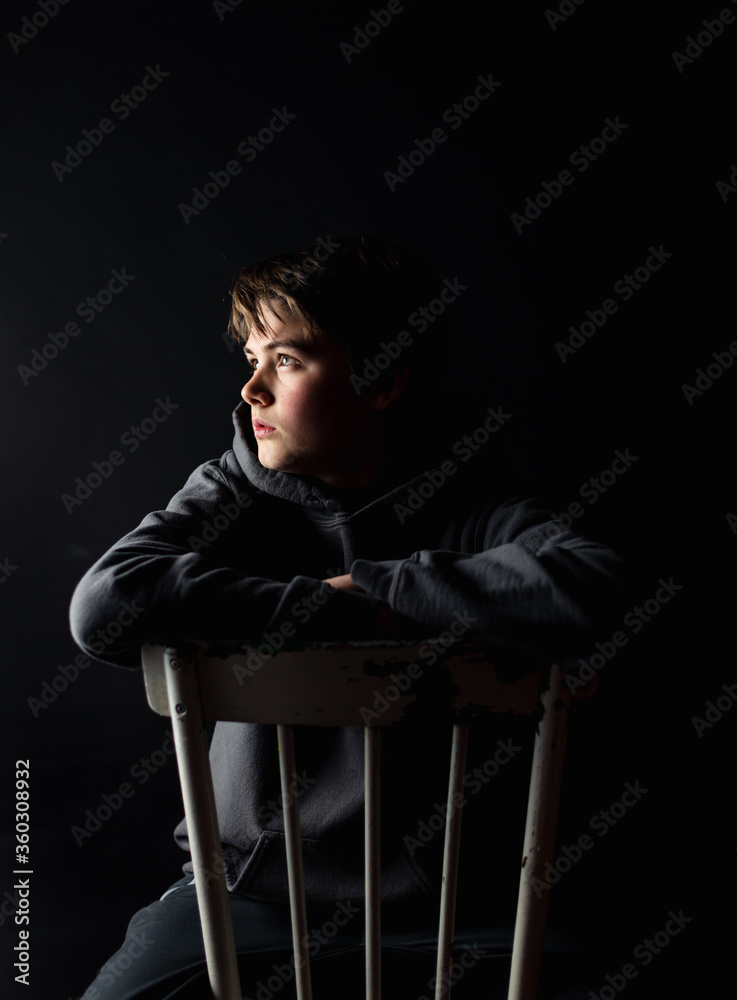 Low key portrait of adolescent boy sitting on a chair in a dark room ...