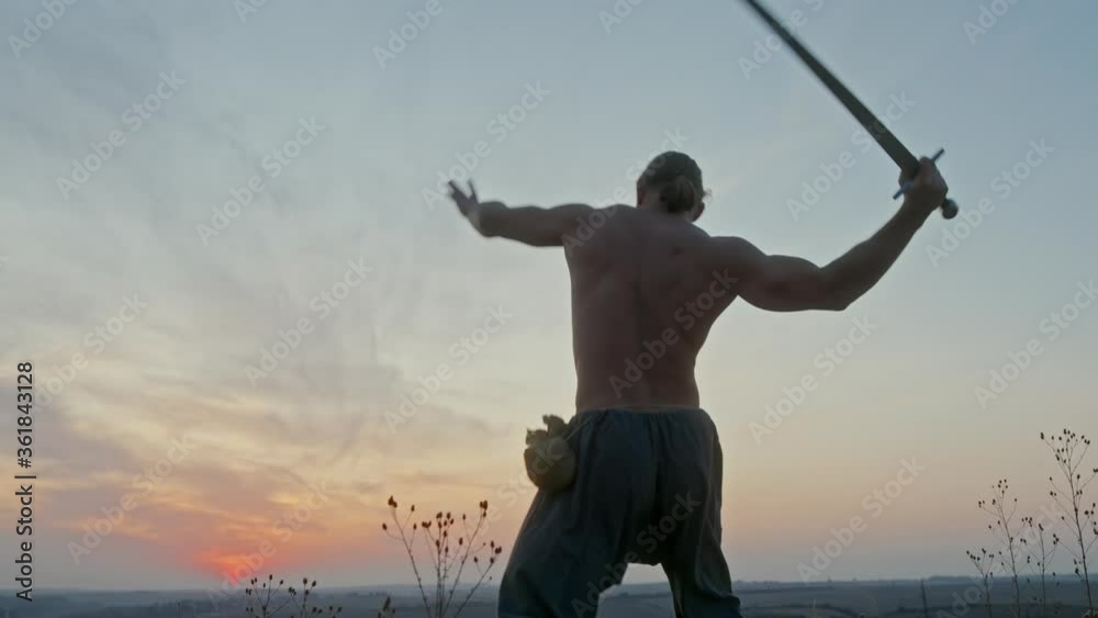Muscular shirtless man with sword practicing slashing movements in a ...