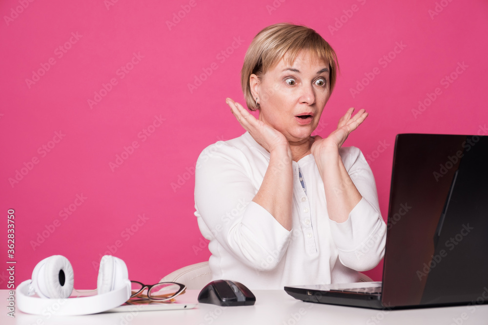 Old woman in her 60s works on computer, wearing headphohes. Laptop on ...