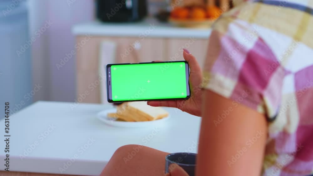 Woman watching a phone with green screen in kitchen while drinking a ...