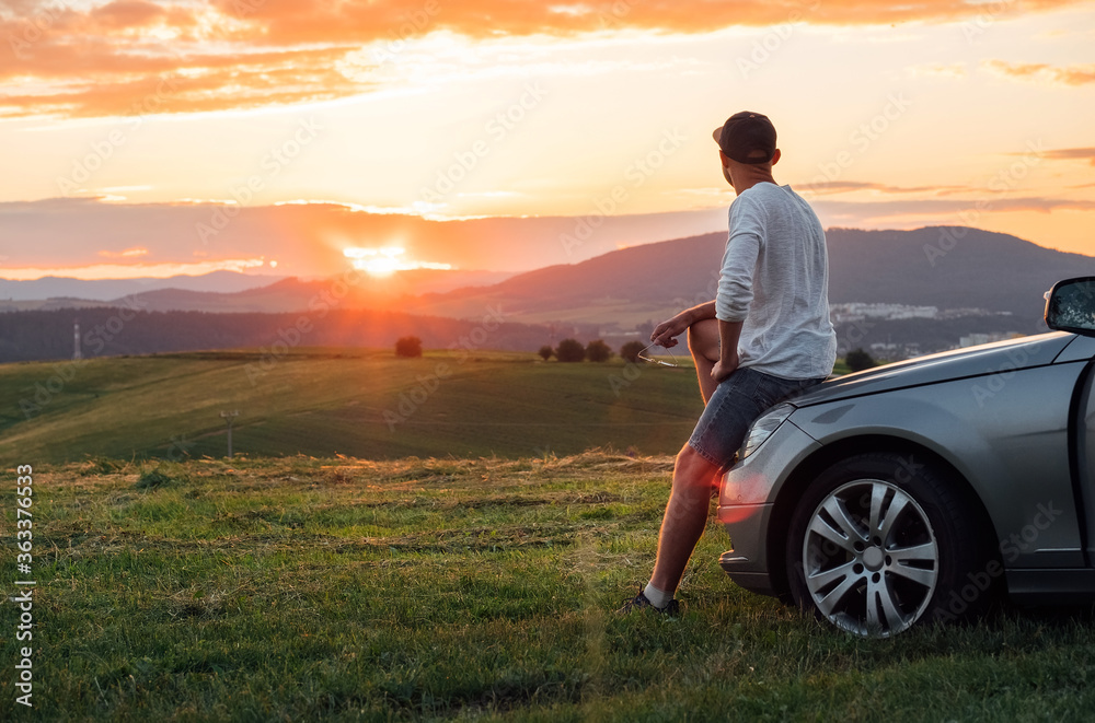 Young man resting during long solo auto trip sitting on his luxury car ...