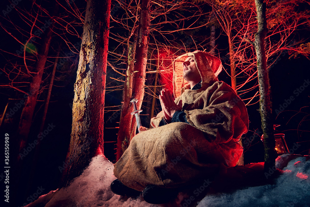 Medieval monk in canvas sackcloth robe praying in dark forest with snow ...