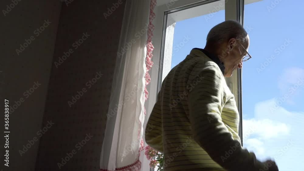 senior man stands at window with open coloured curtains and stretches ...