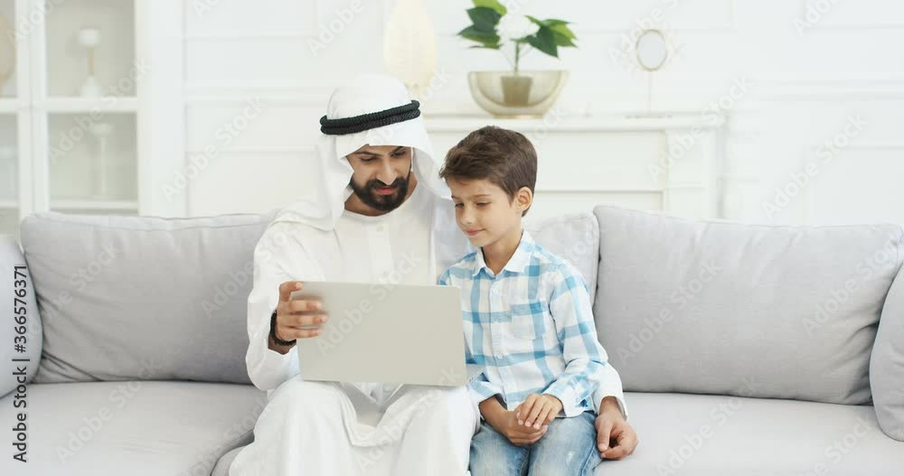 Muslim father in kandura sitting on couch in living room with small ...