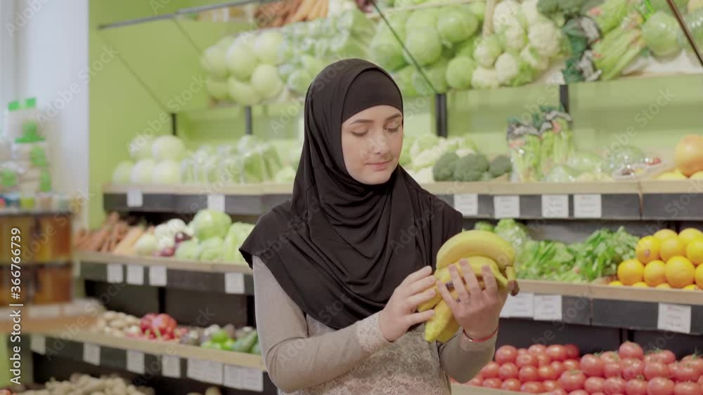 Young Muslim woman examining bunch of bananas in grocery. Portrait of ...