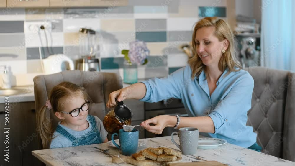 mom pouring tea into cups when sitting at kitchen with child daughter ...