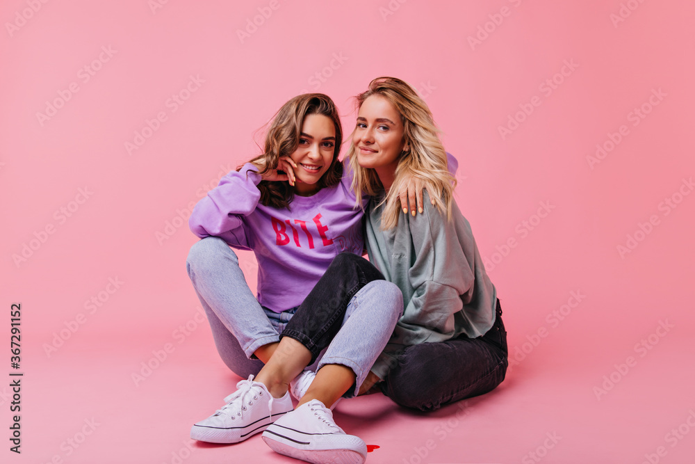 Lovable girls sitting with legs crossed and embracing. Indoor photo of ...