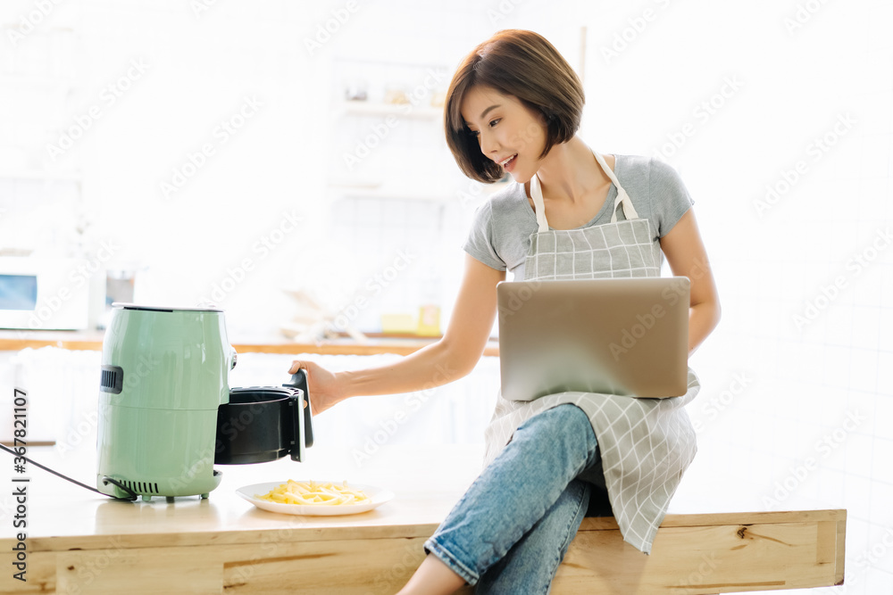 Portrait of beautiful Asian woman working on laptop while cooking with ...