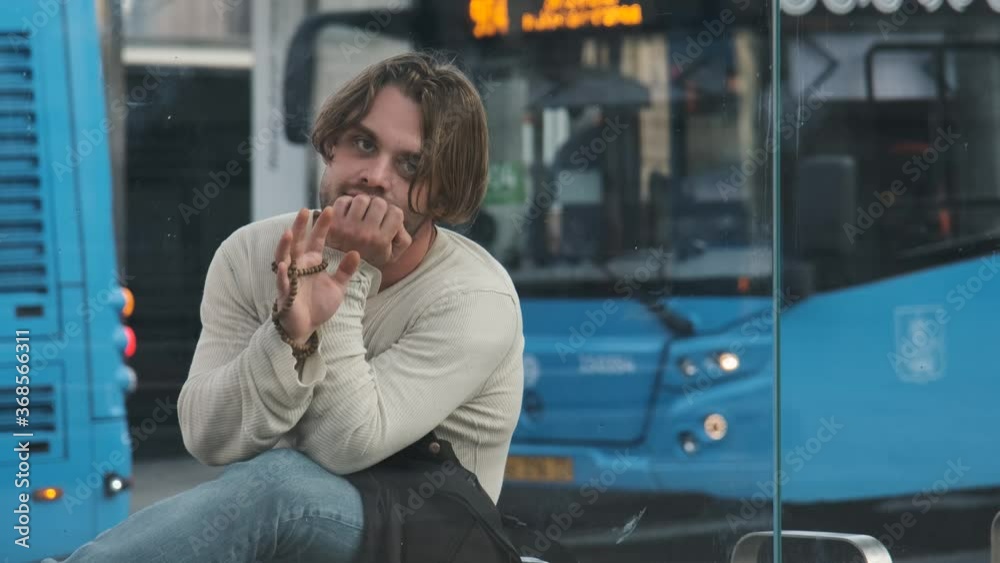 homeless looking hipster sits at bus stop behind the glass in city and ...