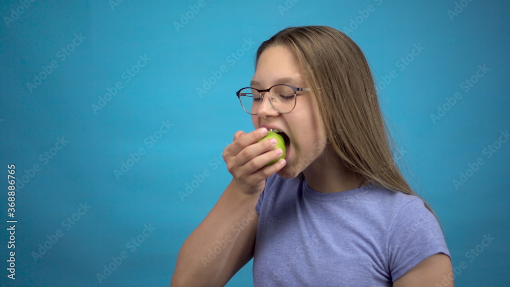 Teenager girl with braces on her teeth eats a green apple on a blue ...