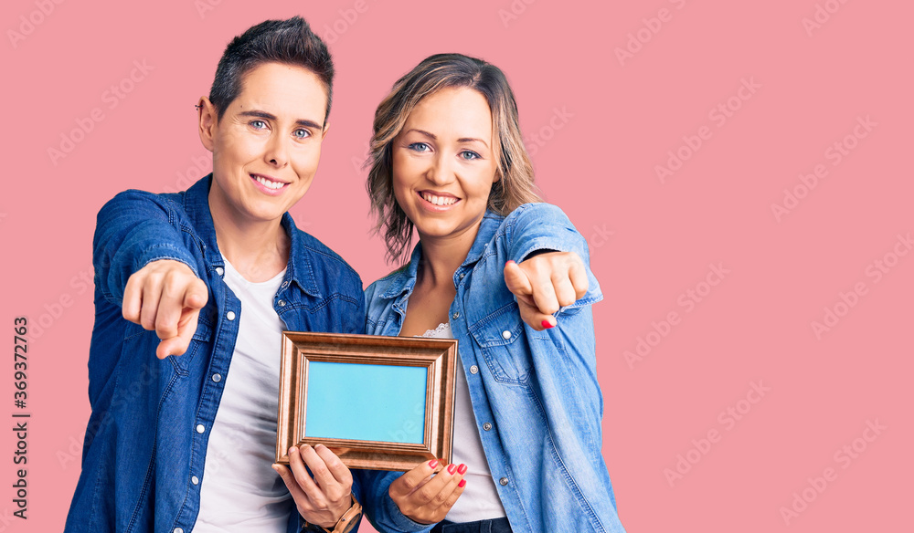 Couple of women holding empty frame pointing to you and the camera with ...