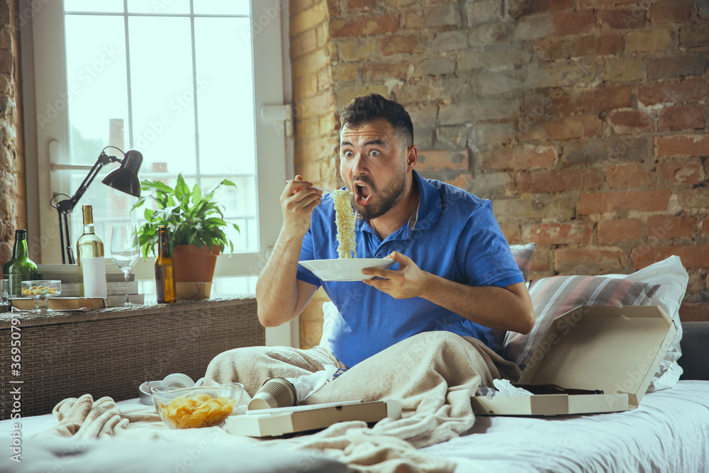 Shocked eating instant noodle. Lazy man living in his bed surrounded ...