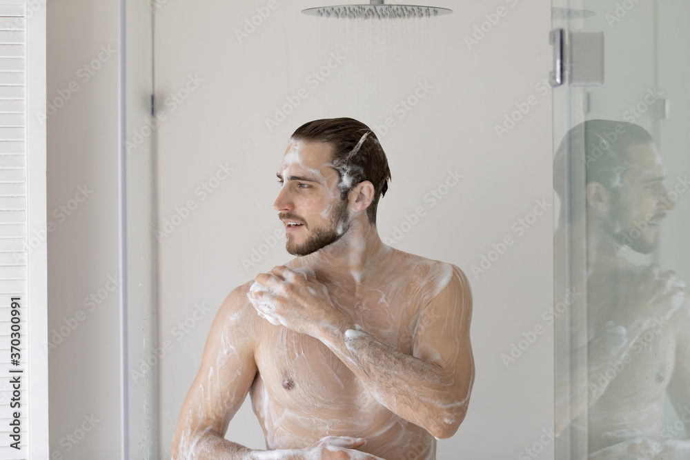 Smiling young man standing in bathroom, taking shower, washing body ...