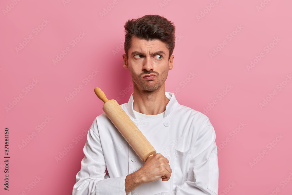 Serious handsome cook looks with gloomy expression aside, holds rolling ...