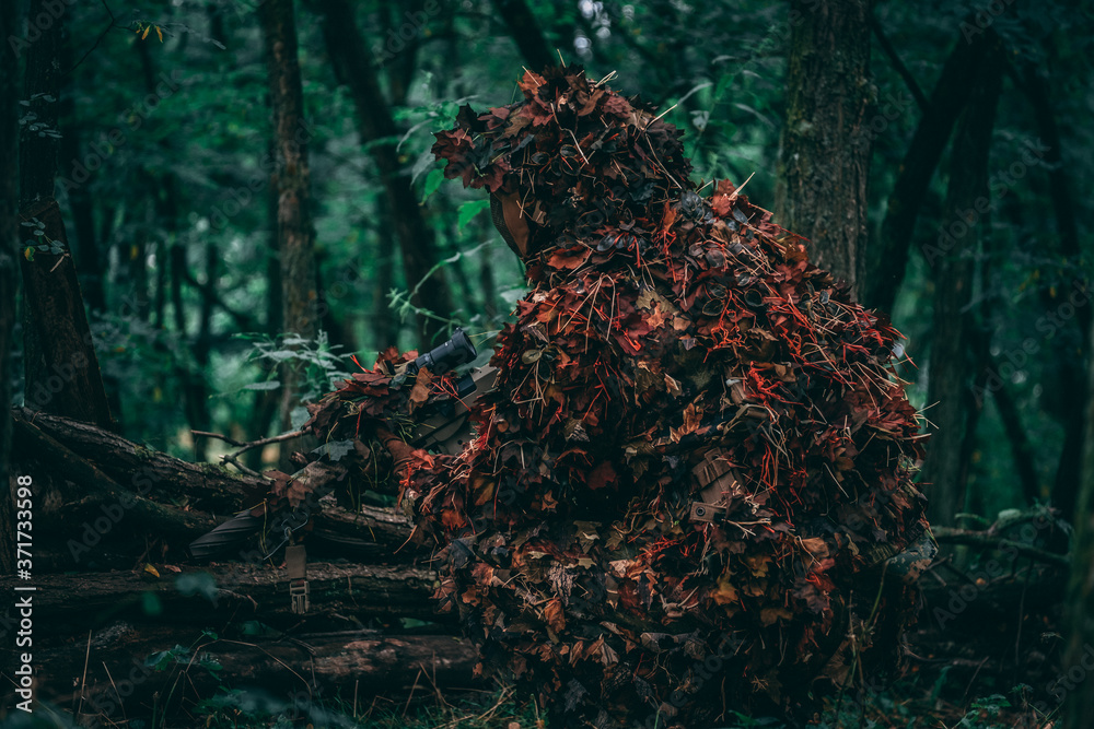 Fully covered sniper in a green forrest. Hiding behind a log. Air-soft ...