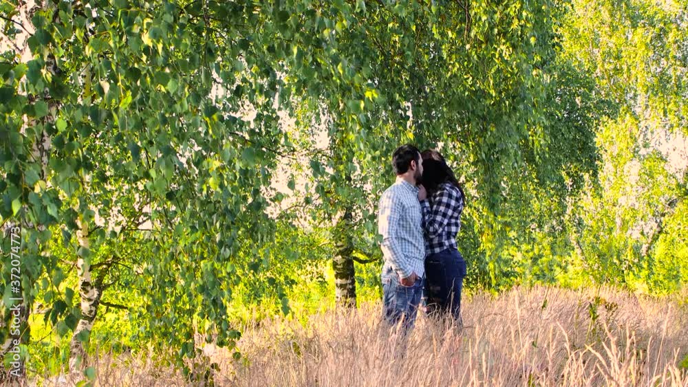 Lovers stand under a tall green birch and hug. The girl gently touches ...