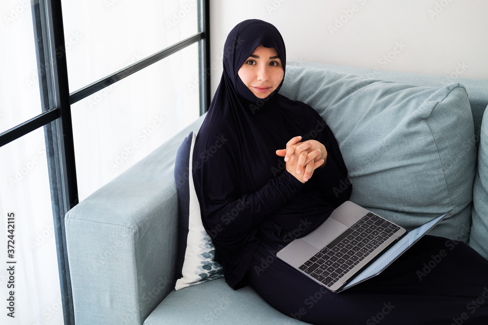 Beautiful young Muslim girl wearing hijab, working at home with laptop ...