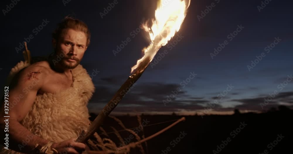 Male hunter in animal hide carrying burning torch and looking at camera ...