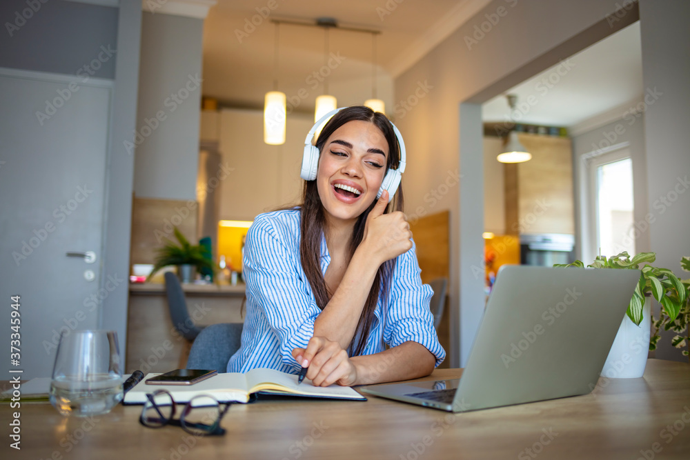 Focused woman wearing headphones using laptop at home, writing notes ...