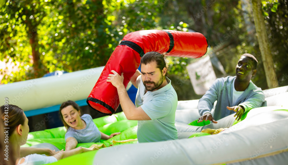 Adults having fun on inflatable amusement playground. Expressive bearded guy fighting off his ...