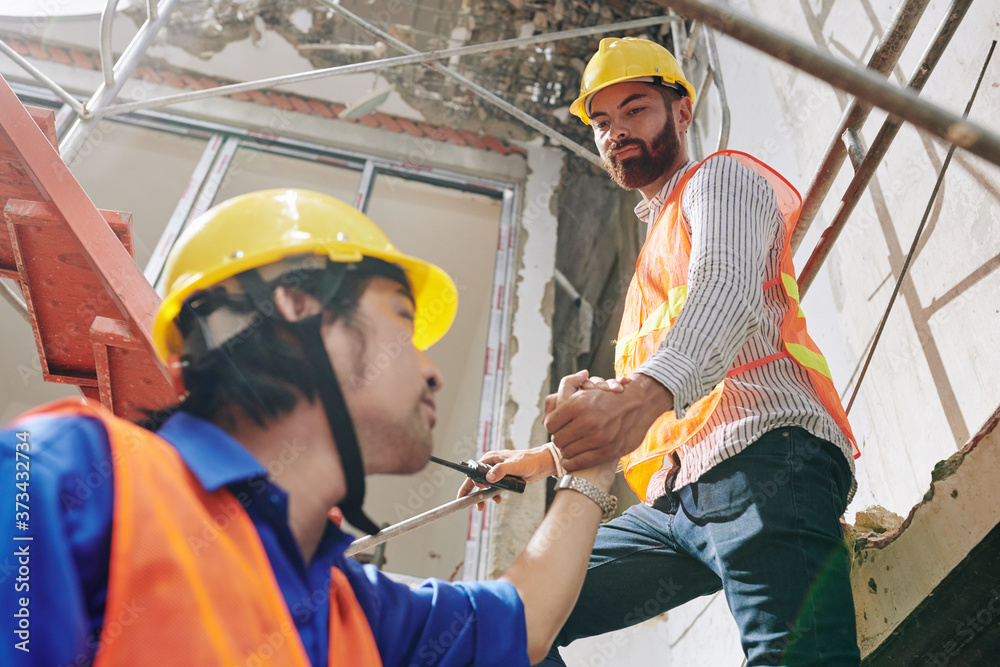 Serious bearded builder helping coworker to climb up the high stairs in ...