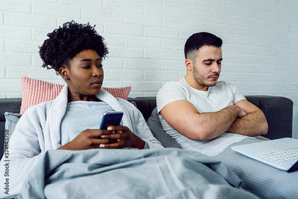 Young multiracial couple using their mobile electronic devices in bed ...