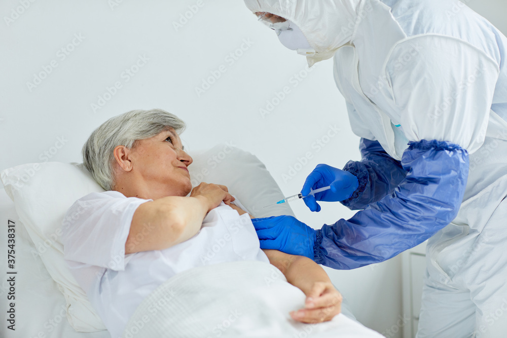 Modern doctor wearing protective suit giving injection to mature woman ...