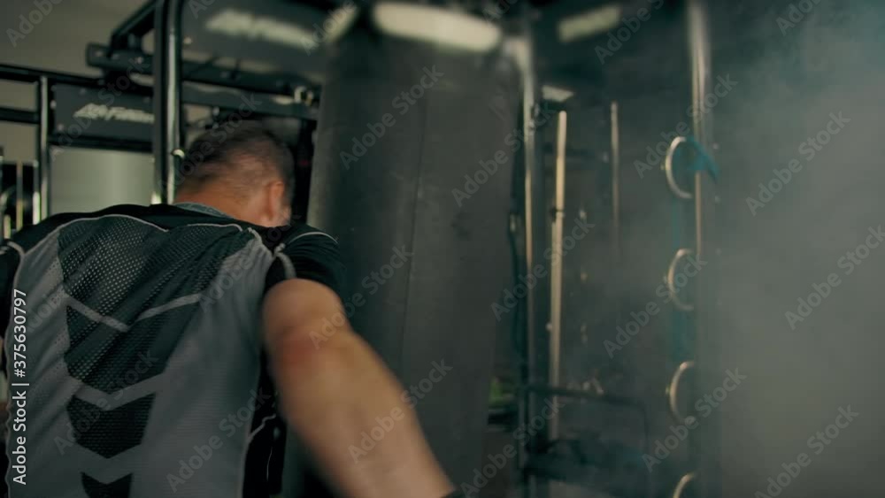 Strong man hitting punching bag indoor of modern gym. Back side view ...