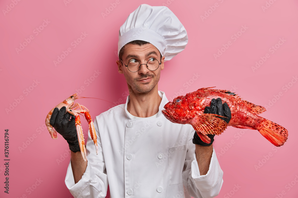 Serious pensive male chef in white uniform, holds crayfish and red sea ...