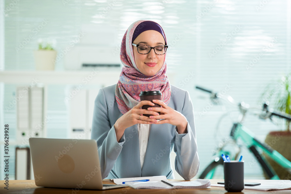 Beautiful young working woman in hijab, suit and eyeglasses sitting in ...