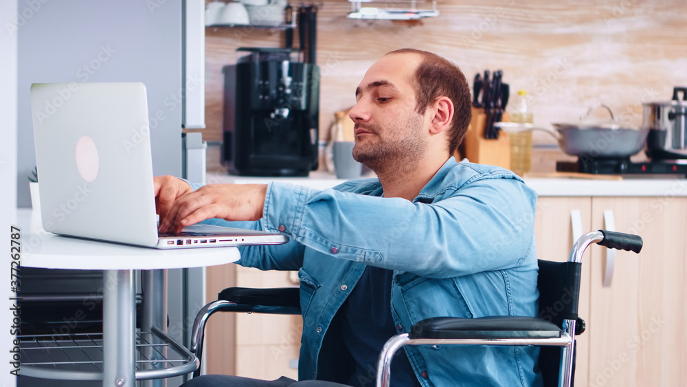 Man with special needs in wheelchair working on laptop in kitchen ...