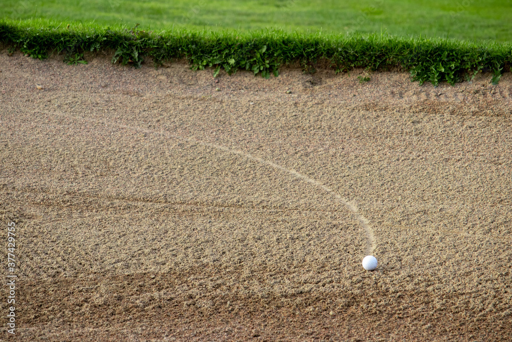 Golf ball lying in a sand bunker with a curved trail visible to show ...
